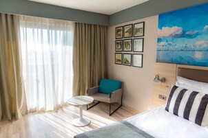 Quarto de hotel com cama, uma cadeira, mesa pequena e parede com quadros.