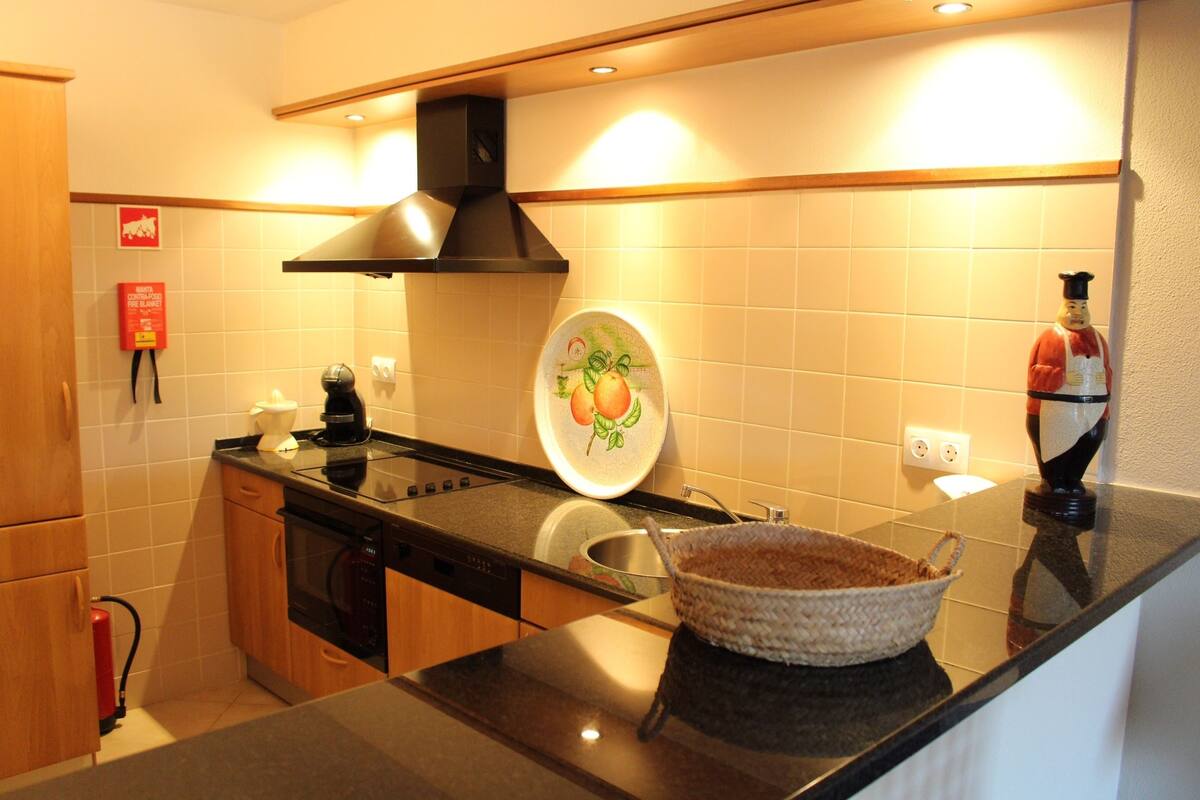Cozinha compacta com fogão, forno e coifa preta. Há um escorredor de louça, uma cafeteira e um prato decorativo com motivos de frutas.