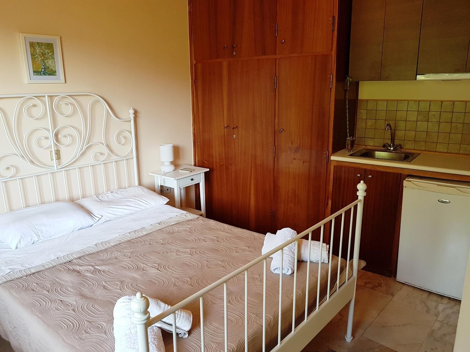 Quarto com cama, mesa de cabeceira, guarda-roupa de madeira e uma pequena cozinha compacta com pia e geladeira.