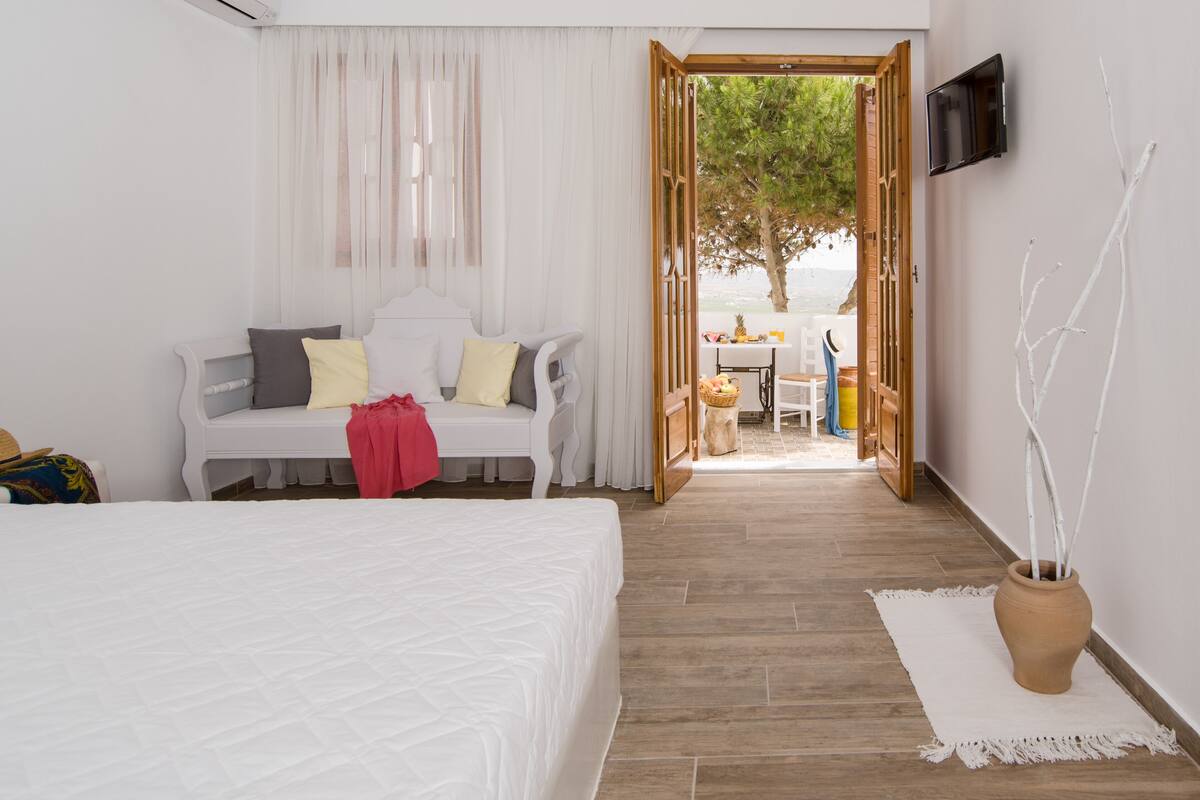 Quarto com cama, sofá branco, televisão e vista para o exterior.