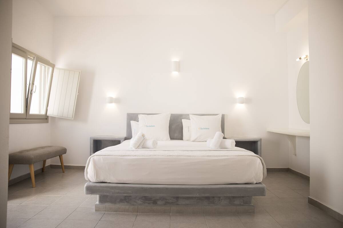 Quarto moderno com uma cama grande, luminárias de cabeceira, um banquinho e um espelho amplo.