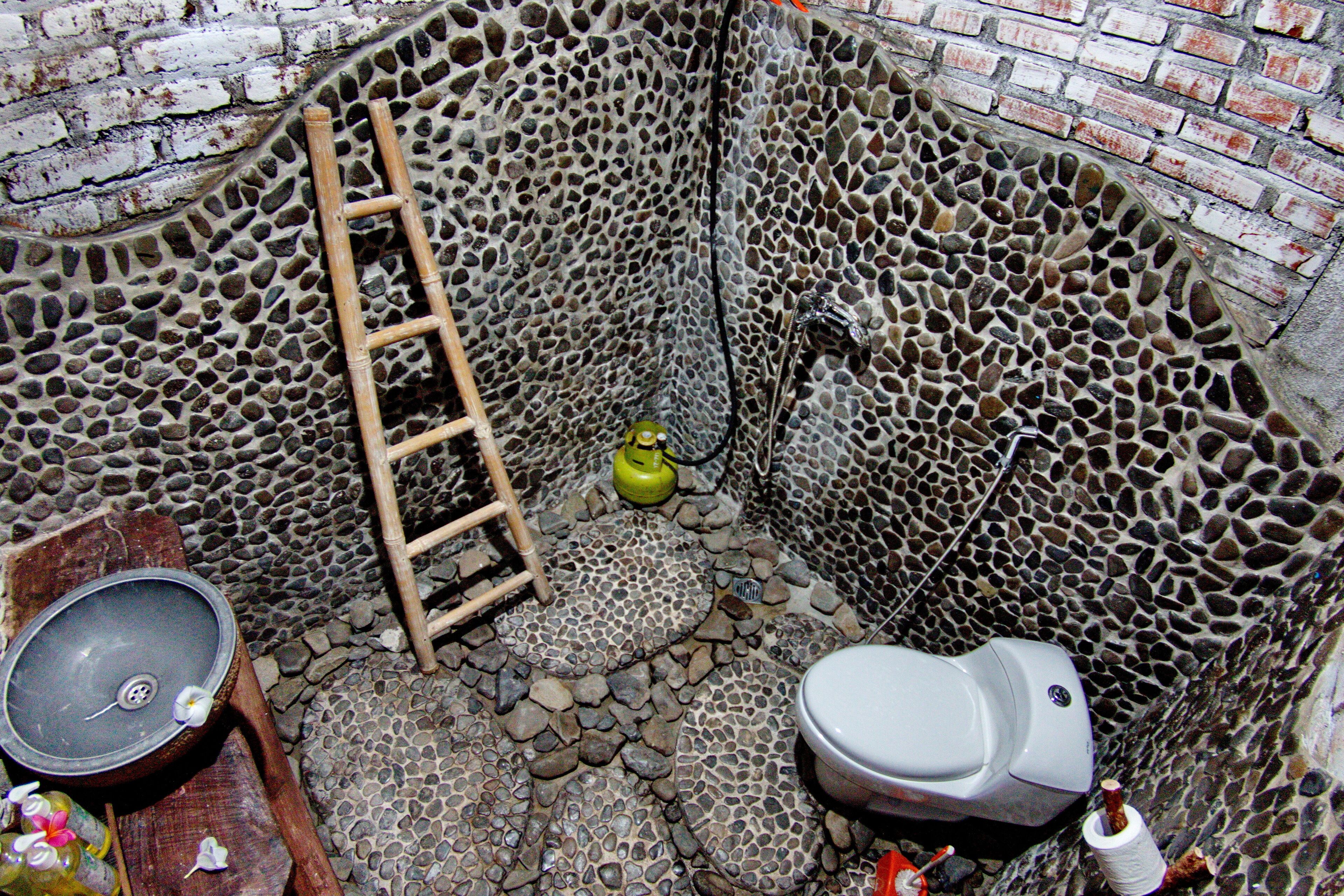 Um banheiro com parede em mosaico de pedra, uma escada de madeira, vaso sanitário e cilindro de gás verde.