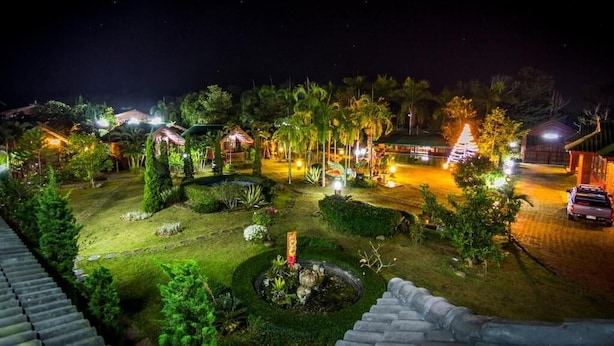 Vista noturna de um jardim bem iluminado, com caminhos, palmeiras e um pequeno lago.