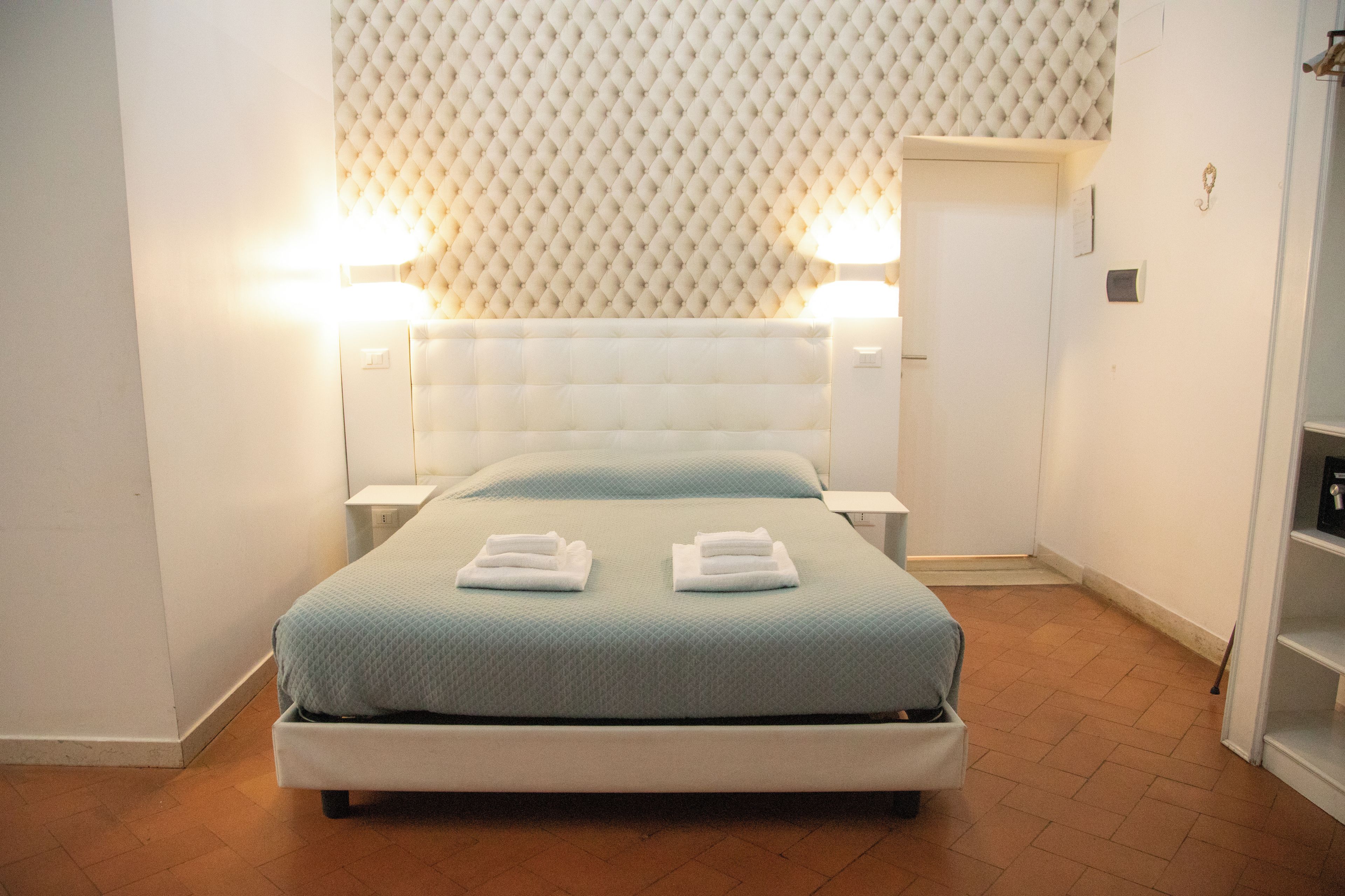 Quarto de hotel com uma cama, duas mesas de cabeceira e uma luminária fixada na parede.