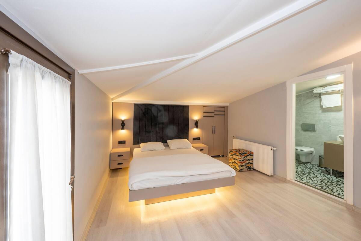 Quarto moderno com cama grande, mesas de cabeceira com abajures, guarda-roupa e banheiro visível por uma porta aberta.