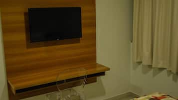 Quarto de hotel com painel de parede de madeira, uma TV de tela plana acima de uma mesa de vidro transparente e uma cama com colcha estampada.