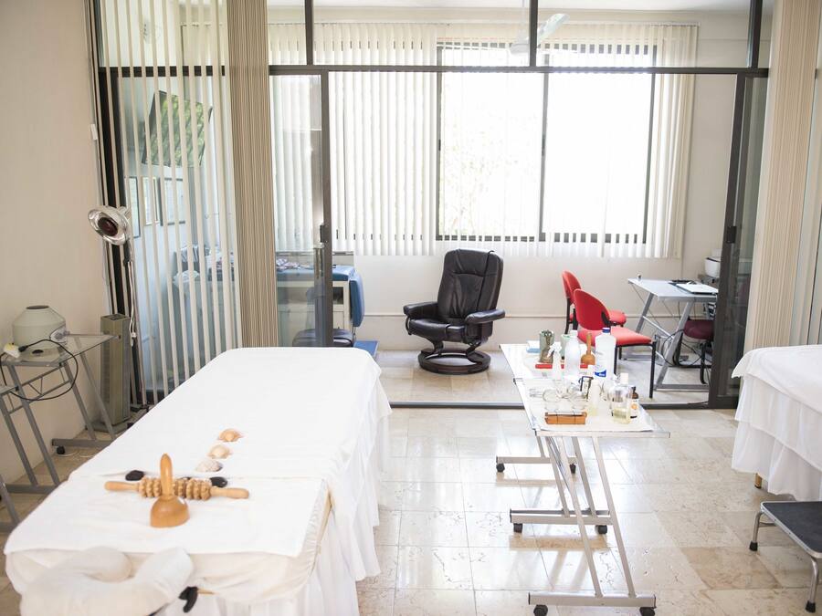Quarto de hotel com cama, mesa de massagem, duas cadeiras, escrivaninha e janela com persianas.