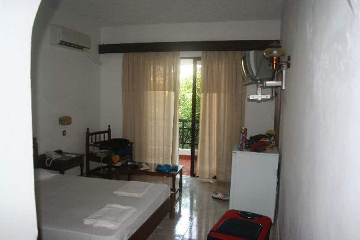 Quarto pequeno e compacto com uma cama, uma escrivaninha e uma cadeira. Há uma janela com cortinas, um radiador e uma mesa pequena com objetos sobre ela.