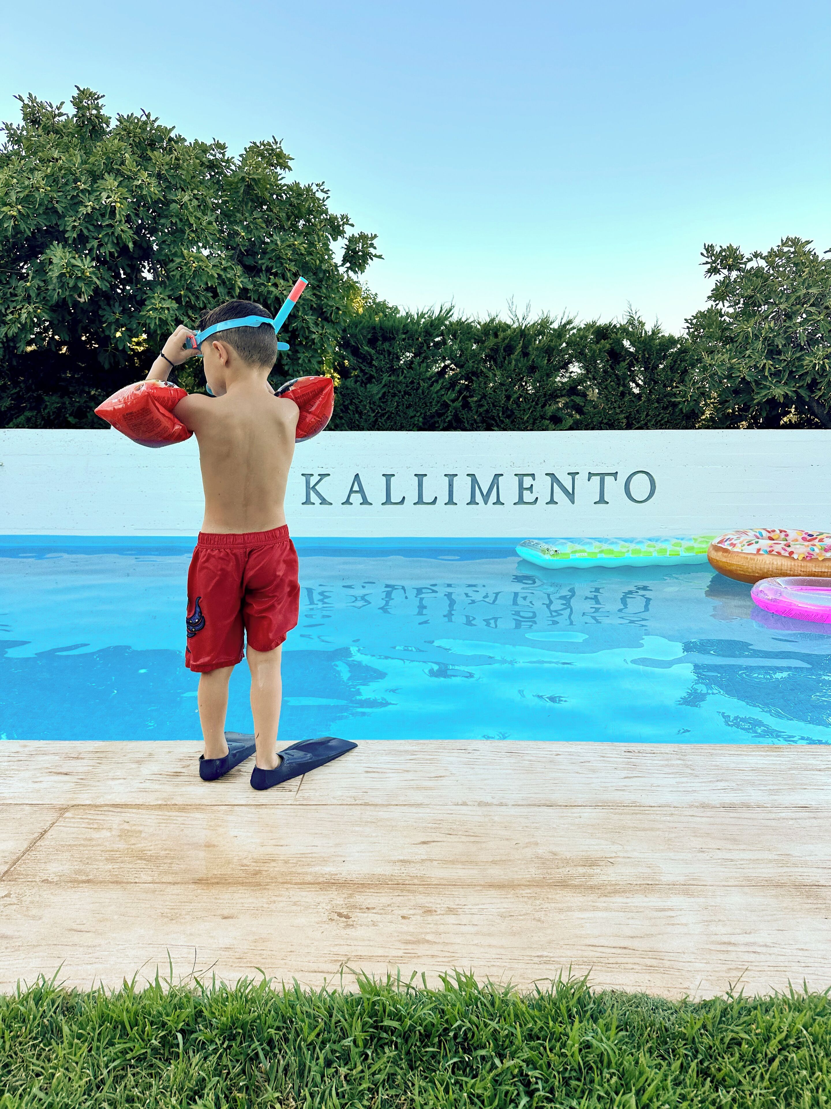 Criança com sunga vermelha e touca azul parada perto de uma piscina com uma placa que diz 'KALLIMENTO'.
