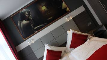 Quarto de hotel moderno com uma cama grande, roupa de cama vermelha e branca, televisão fixada na parede e cabeceira escura.