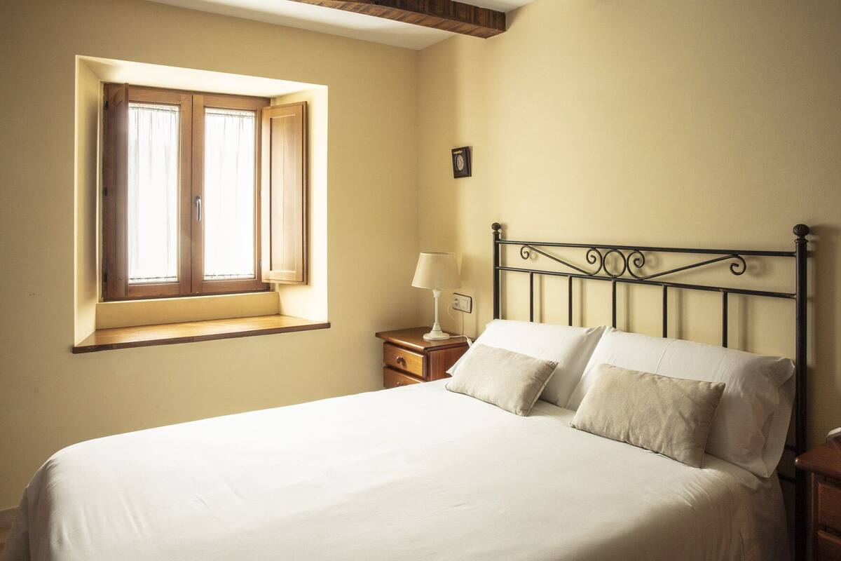Quarto com cama, mesa de cabeceira, abajur e janela com cortinas.