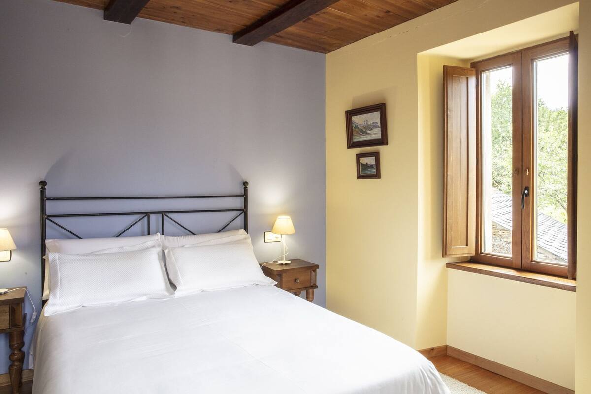 Quarto com uma cama, duas luminárias de cabeceira, janela com vista e duas quadros na parede.