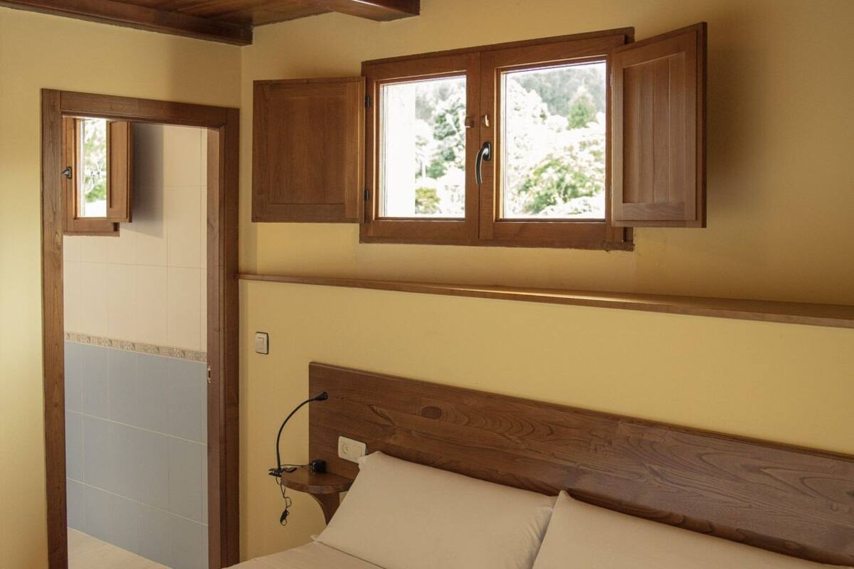 Quarto com teto de madeira, cama com roupa de cama branca, mesa de cabeceira e janela com vista para o verde.