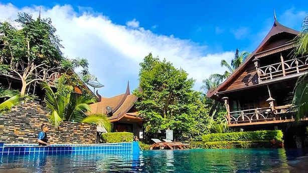 Uma piscina cristalina com uma parede de pedra e um deck de madeira de um lado, cercada por vegetação exuberante e arquitetura tradicional.