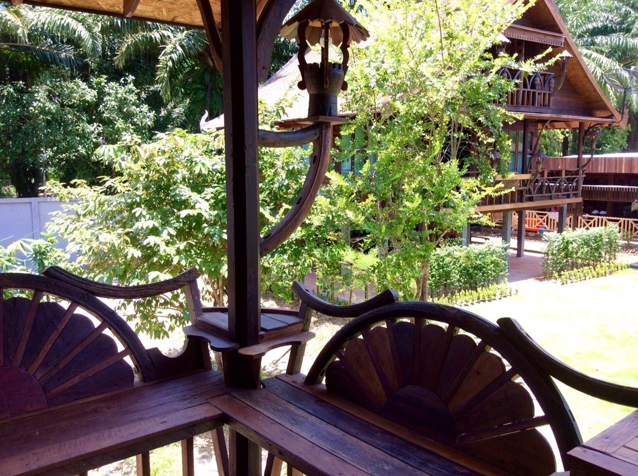 Um deck de madeira com estrutura de gazebo, cercado por vegetação exuberante e um pequeno edifício ao fundo.