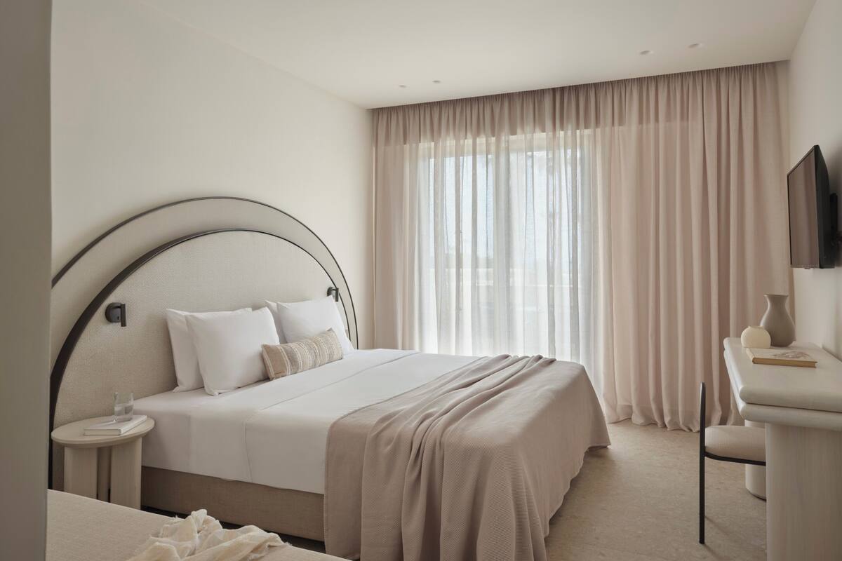 Quarto de hotel com uma cama, uma mesa de cabeceira com um copo de água, uma cadeira e uma televisão na parede.
