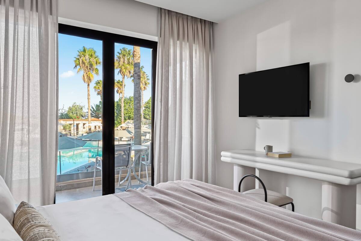 Quarto de hotel com uma cama, uma televisão na parede, uma mesinha com um livro e uma chávena, e vista para uma piscina e palmeiras pela janela.