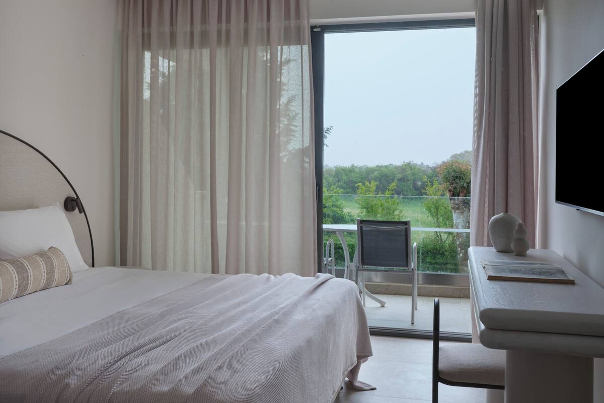 Quarto de hotel com cama, escrivaninha, cadeira, televisão e varanda com vista para o verde.
