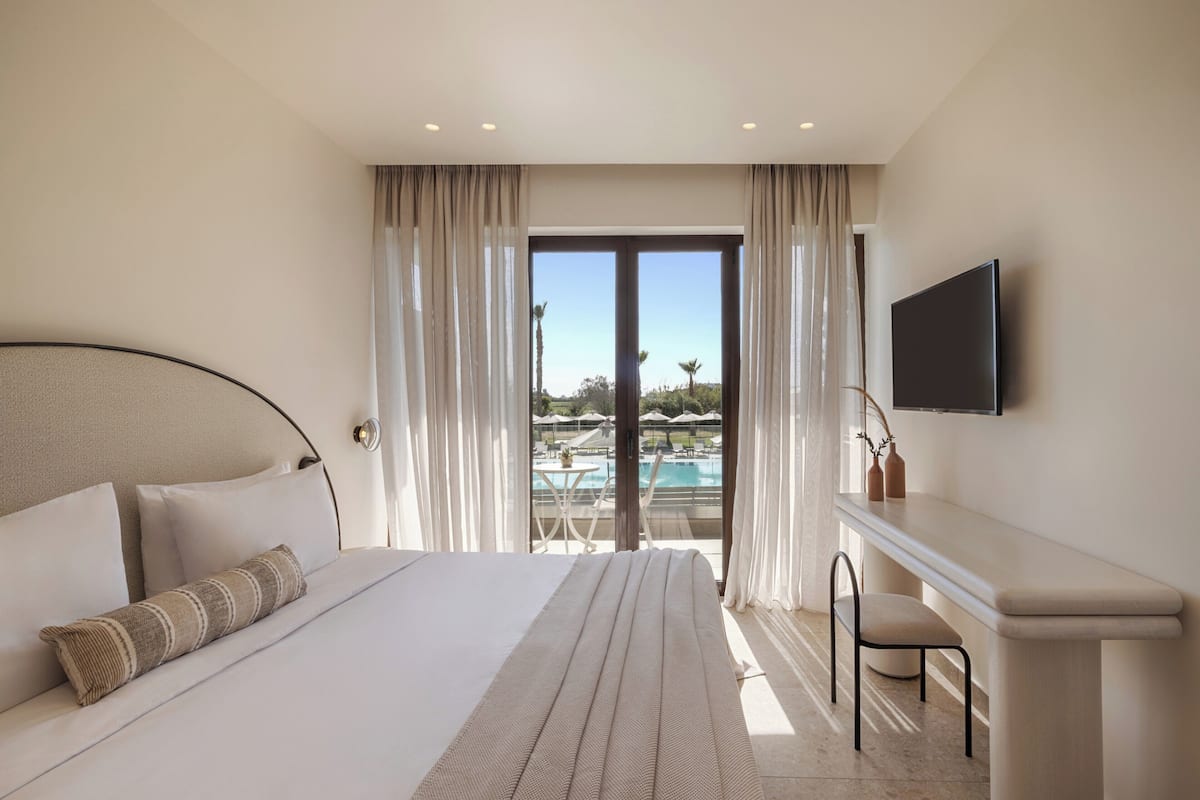 Quarto de hotel com cama, televisão, escrivaninha e vista para a piscina.