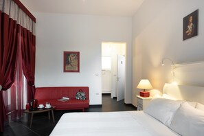 Quarto de hotel com uma cama, um sofá vermelho, uma mesa pequena com cafeteira, uma mesinha de cabeceira com abajur e uma porta para o banheiro.