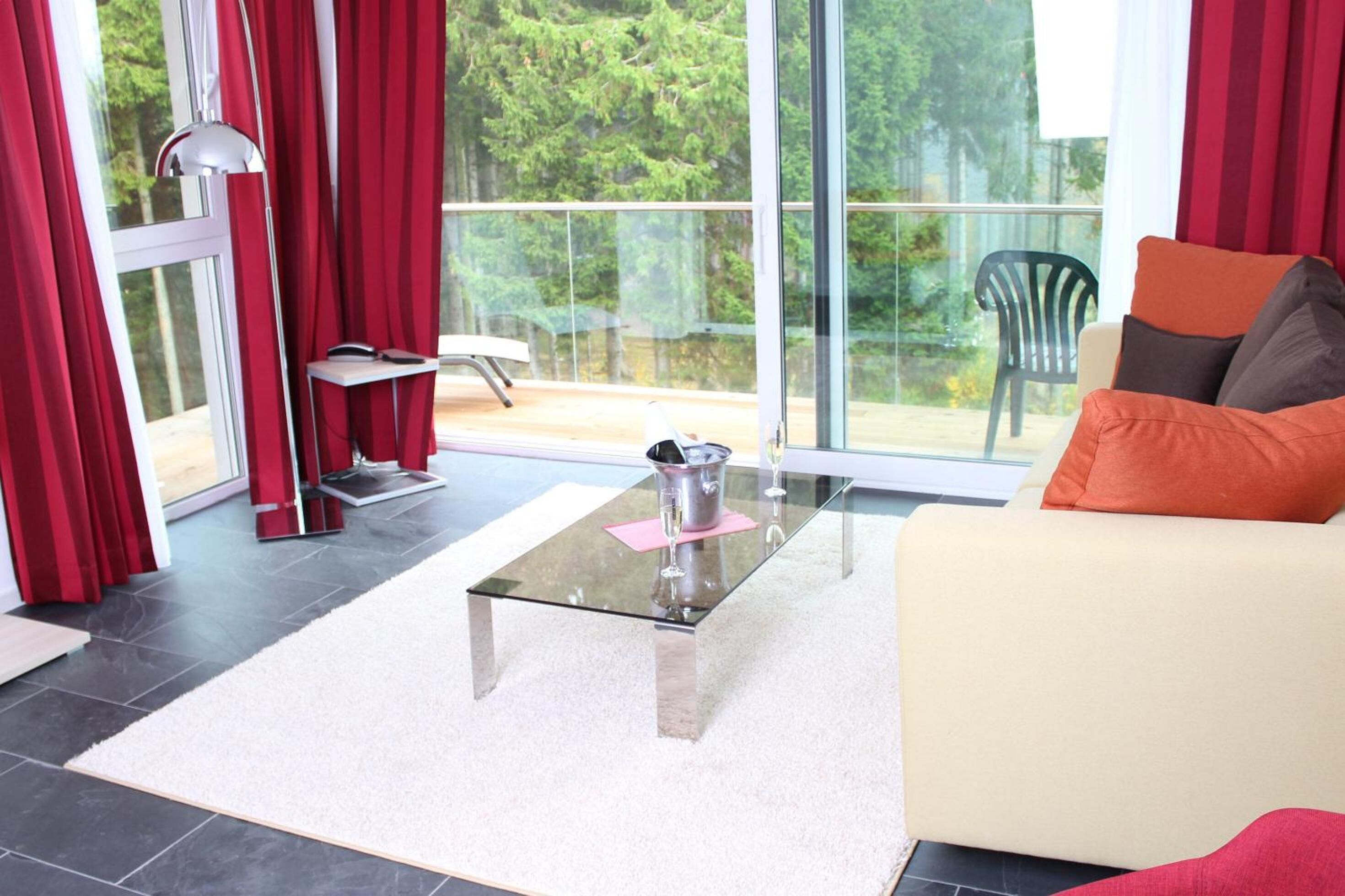 Sala de estar moderna com mesa de centro de vidro, sofá com almofadas laranja e vista para uma varanda com cadeira.