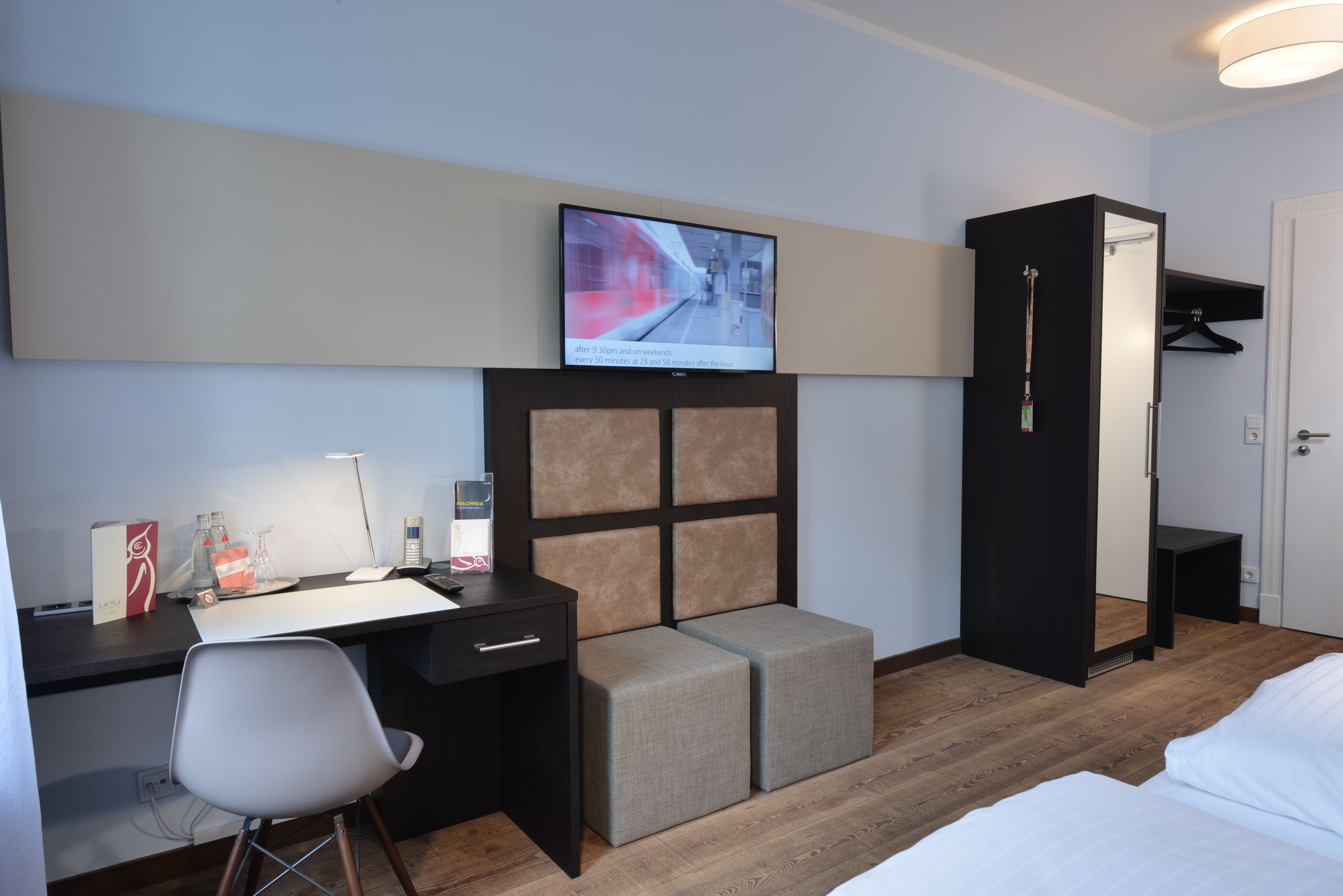 Quarto de hotel moderno com escrivaninha, cadeira, dois pufes, cama, televisão na parede e guarda-roupa.