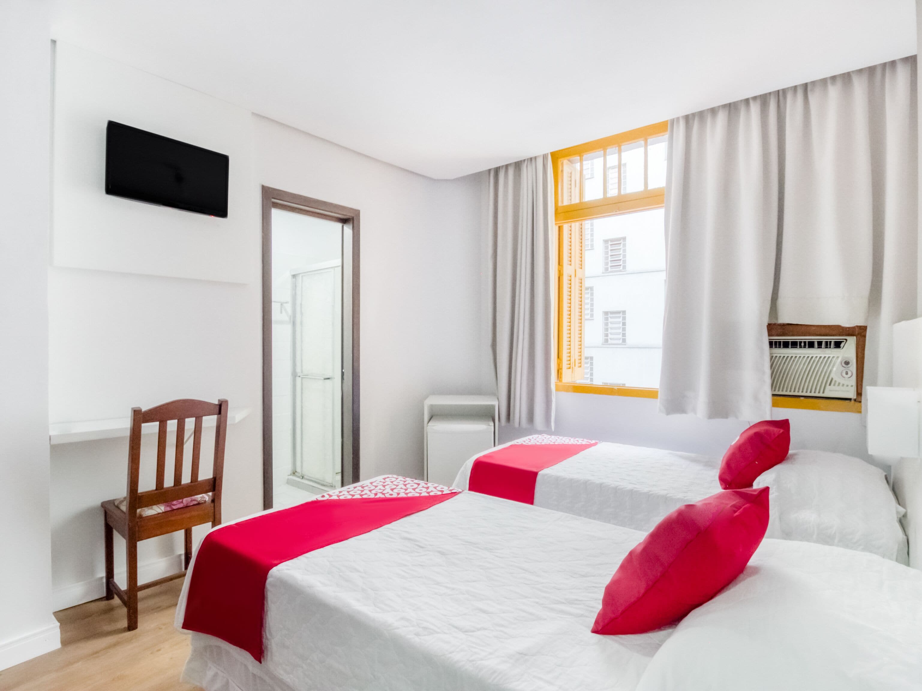 Quarto de hotel com duas camas, uma cadeira de madeira, uma escrivaninha, televisão, janela com cortinas e radiador.