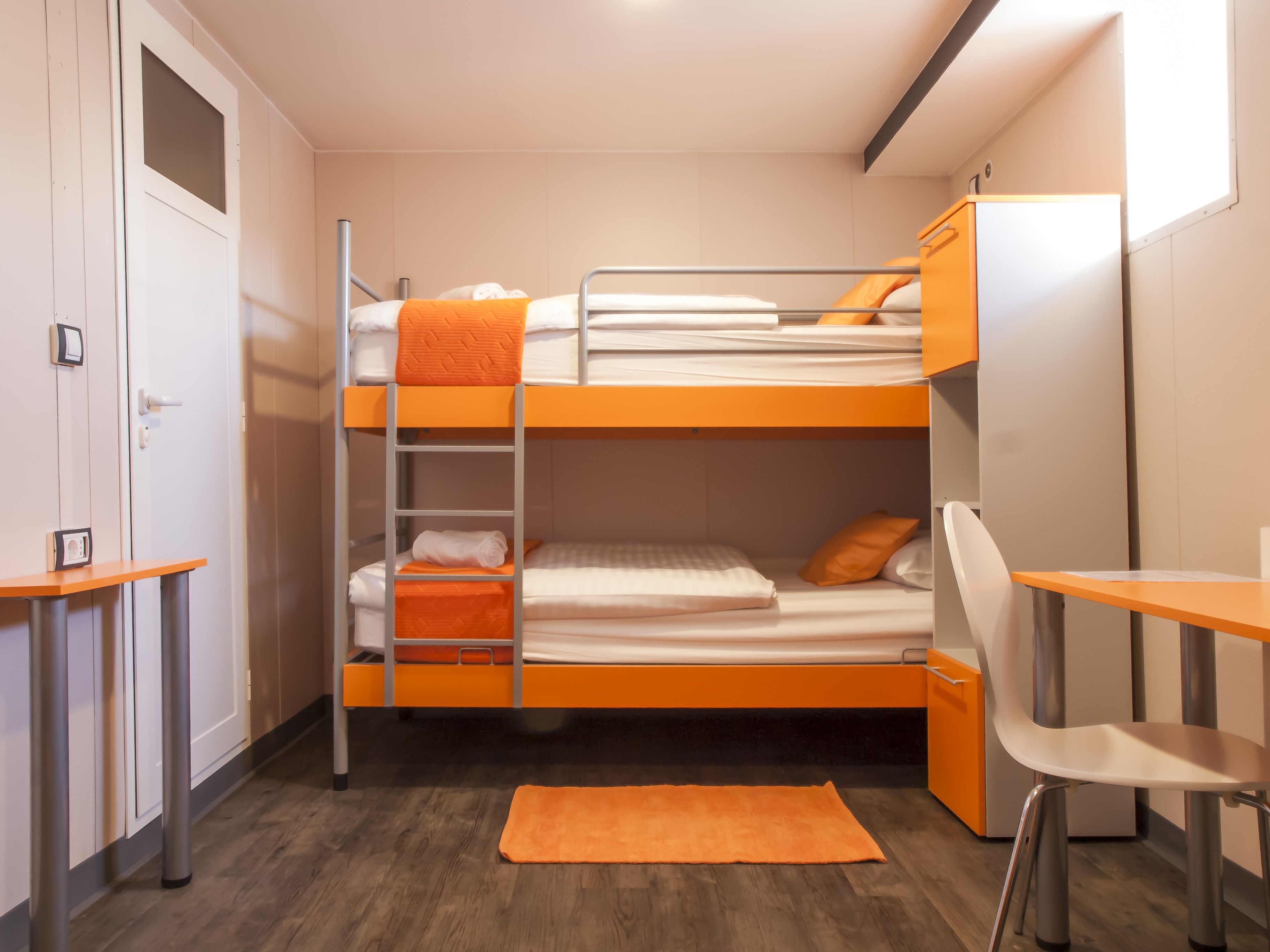 Quarto com beliche, escrivaninha e cadeira, um tapete pequeno e uma porta com fechadura.