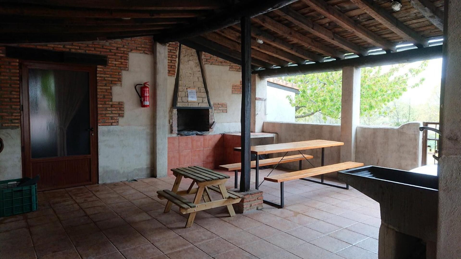 Um pátio externo com parede de tijolos, um extintor de incêndio, um banco de madeira, mesas de piquenique e uma churrasqueira.