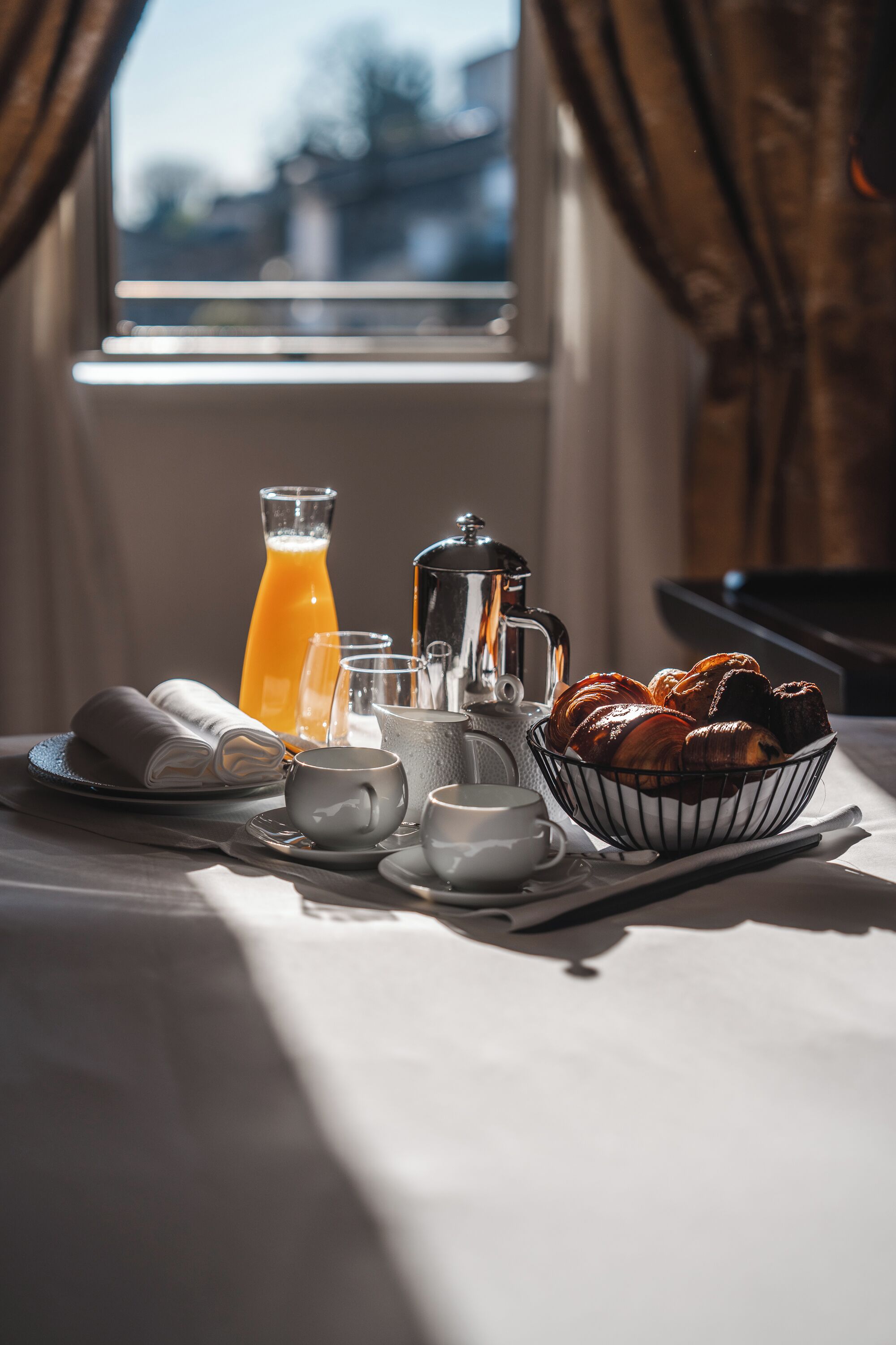 Uma mesa perto de uma janela com disposição para o café da manhã: sumos de laranja, jarra de café, croissants e uma travessa de pastelaria.