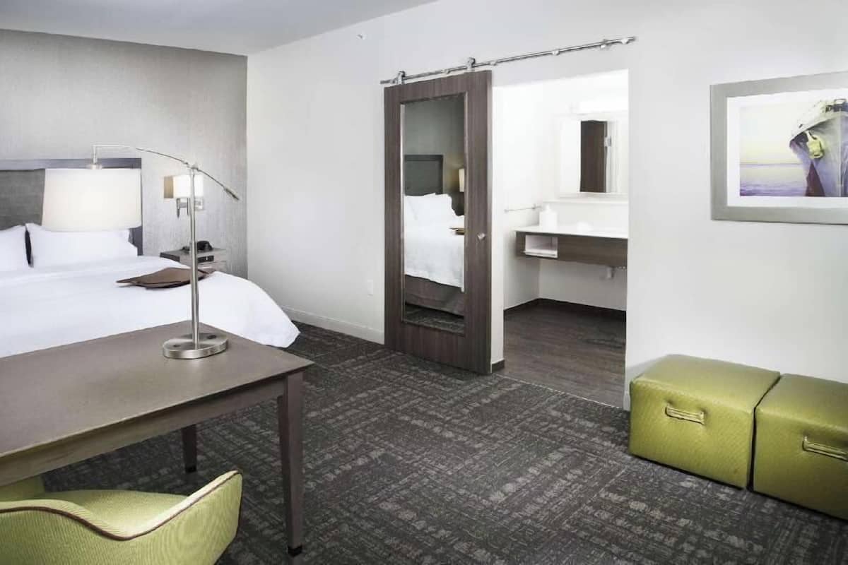 Quarto de hotel com cama, escrivaninha, cadeira, banheiro com espelho e um armário verde.