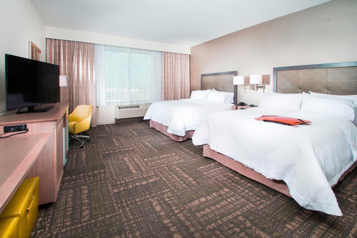 Quarto de hotel com duas camas, uma mesa de trabalho com televisão, uma cadeira e janela com cortinas.