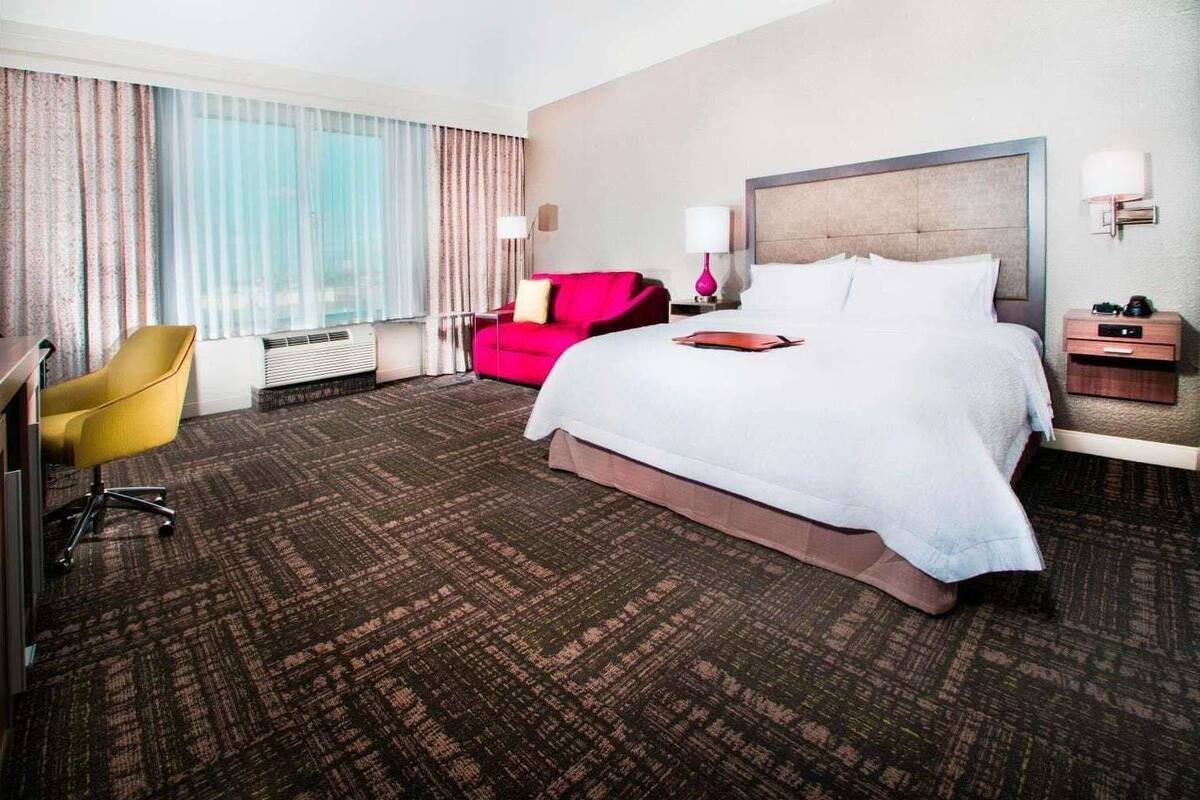 Quarto de hotel com uma cama grande, uma escrivaninha, uma cadeira e uma janela com cortinas.