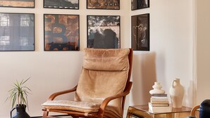 Um quarto com uma cadeira de madeira, um banco estofado, uma pequena mesa com livros e vasos, e uma planta em vaso.
