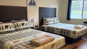 Quarto de hotel com duas camas, cada uma com colcha e cabeceira com tema de floresta. Há uma janela com vista para o exterior, uma mesa de cabeceira com abajur e um quadro pendurado na parede.