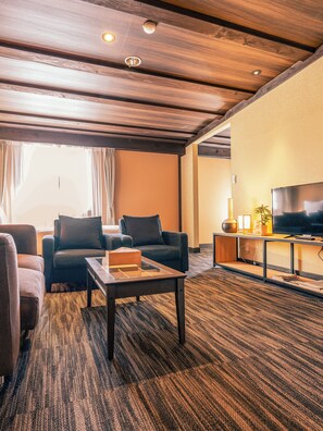 Quarto de hotel com teto de madeira, TV de tela plana, sofá, mesa de centro e piso coberto por carpete.