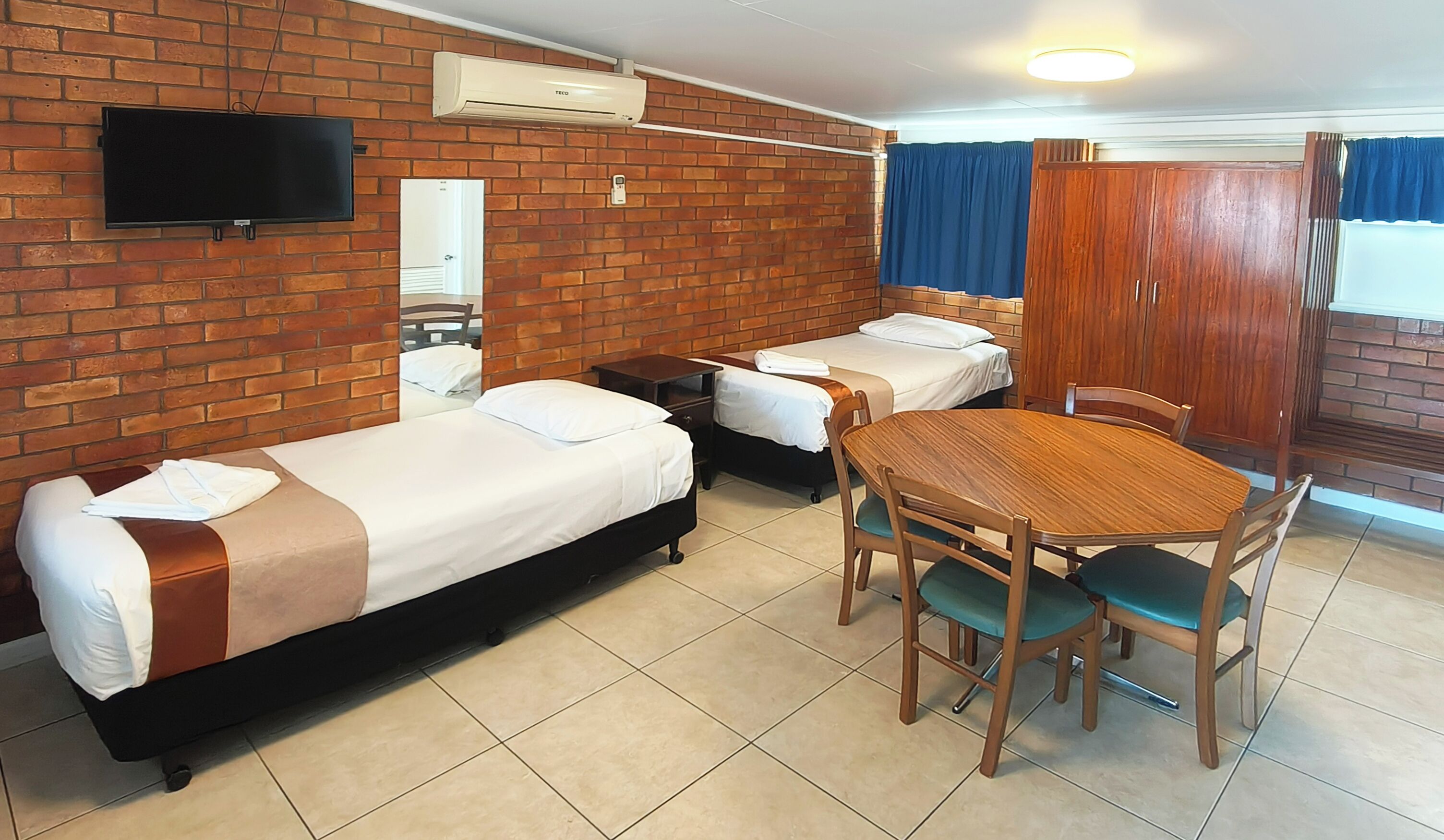 Quarto de hotel com duas camas de solteiro, uma mesa de jantar de madeira com cadeiras, uma TV fixada na parede de tijolos e um aparelho de ar condicionado.