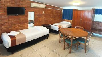 Quarto de hotel com duas camas de solteiro, uma mesa de jantar de madeira com cadeiras, uma TV fixada na parede de tijolos e um aparelho de ar condicionado.