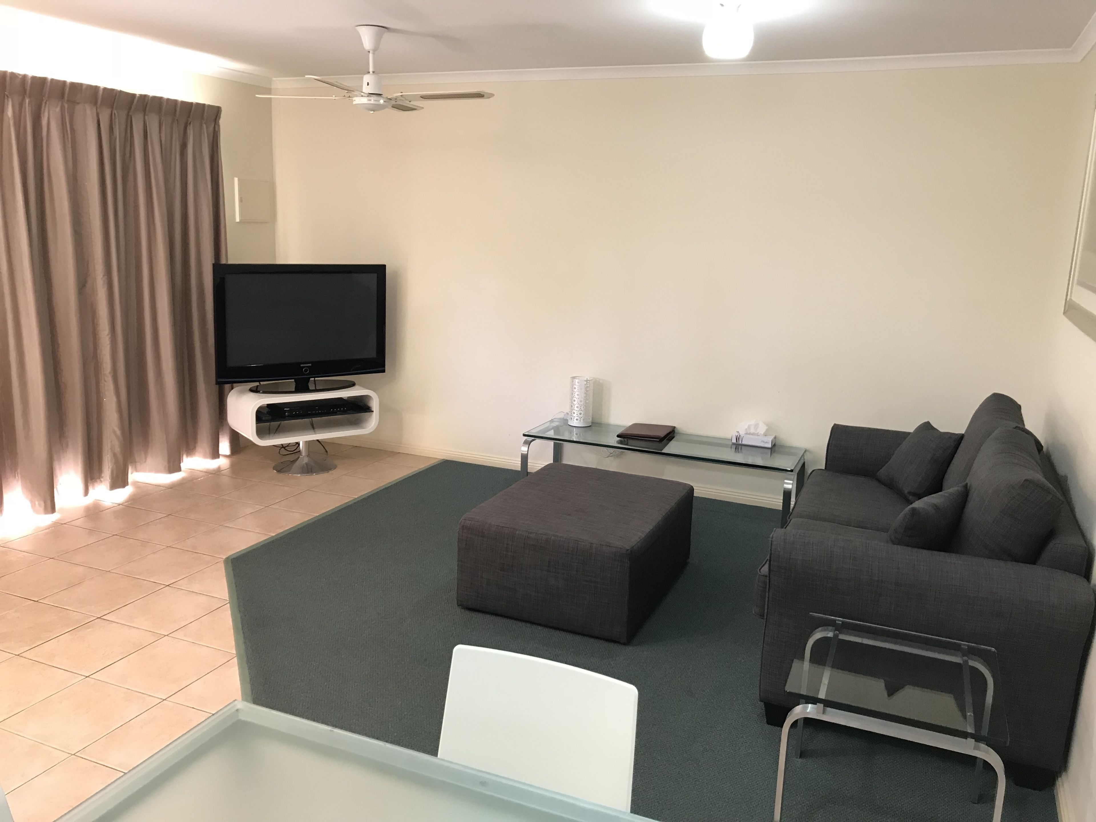Uma sala de estar com televisão de tela plana, um sofá, uma mesa de centro de vidro e um ventilador de teto.