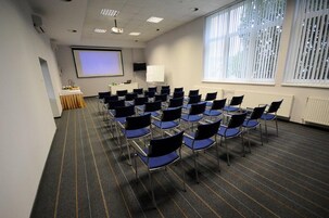 Uma sala de conferência com fileiras de cadeiras, uma tela de projetor, uma lousa e uma janela com persianas.