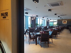 Uma sala de jantar espaçosa com várias mesas e cadeiras, janelas amplas e iluminação no teto.