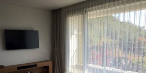 Quarto com televisão de tela plana na parede, janela com cortinas translúcidas e vista para árvores e folhagem lá fora.