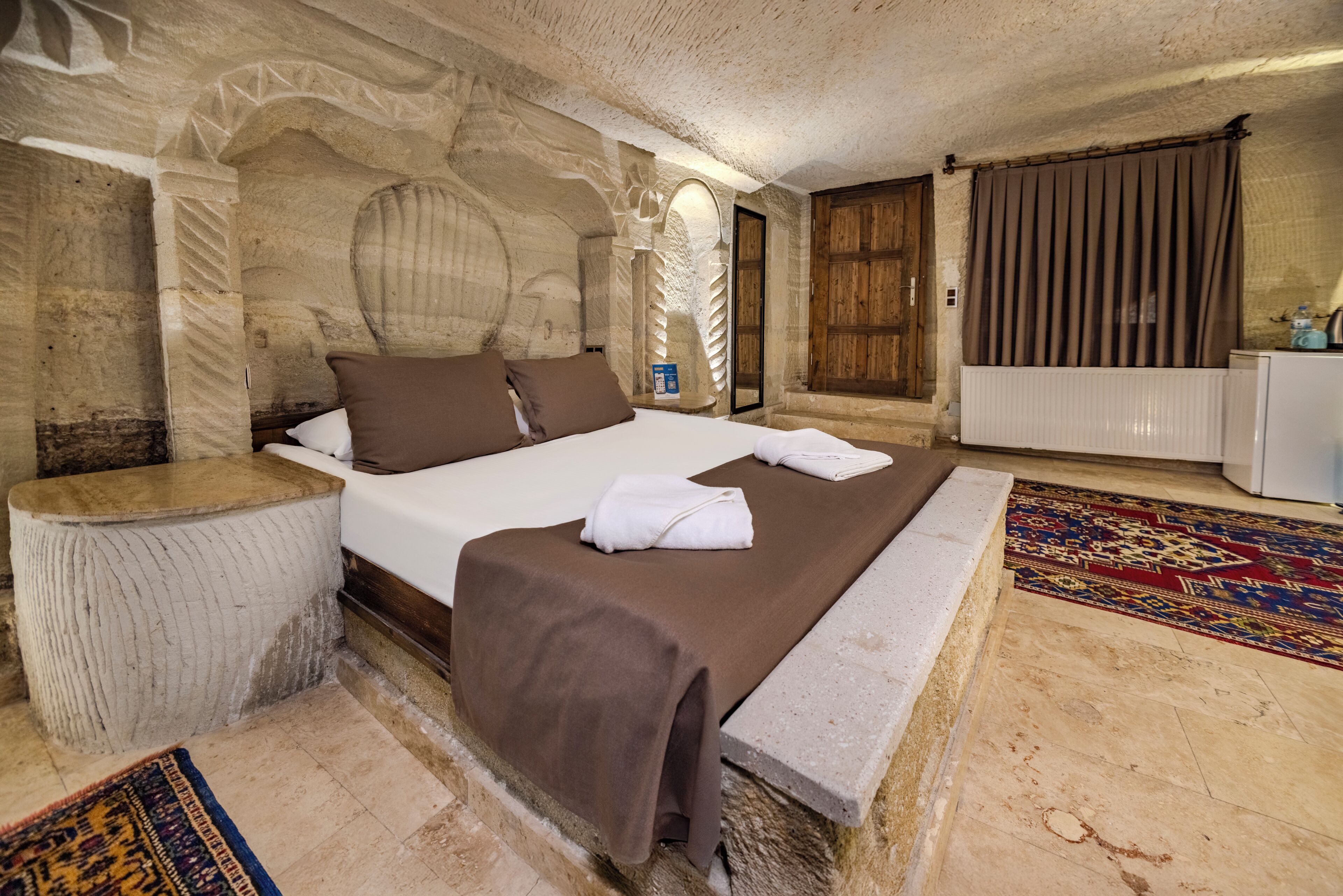 Um quarto com cama, mesa de cabeceira de madeira, parede de pedra, porta de madeira e tapete estampado.