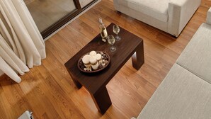 Uma sala de estar moderna com uma grande porta de vidro deslizante que dá para uma varanda. O ambiente possui um sofá branco, uma mesa de centro de madeira com uma tigela decorativa e uma pequena mesa lateral.