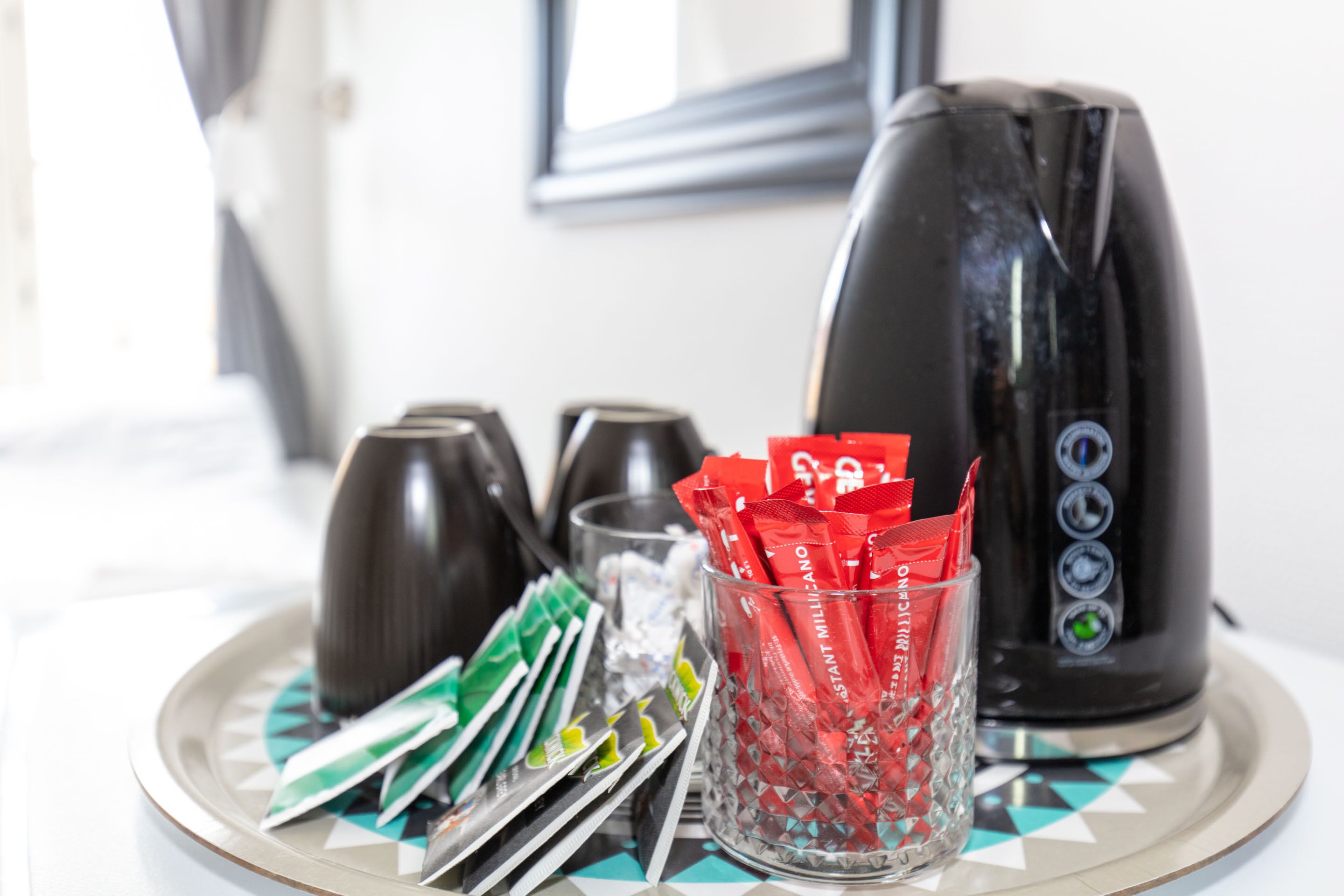 Uma cafeteira, um copo com sachês vermelhos e uma bandeja com sachês verdes e brancos sobre uma mesa.