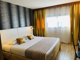 Quarto de hotel com uma cama grande, cabeceira com design texturizado e vista da cidade através de cortinas leves.