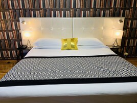 Cama bem arrumada com colcha estampada, um travesseiro amarelo e duas luminárias laterais. Uma estante cheia de livros aparece ao fundo.