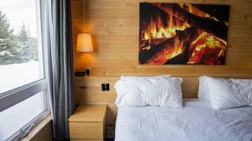 Quarto de hotel com cama, mesa de cabeceira, luminária, quadro de fogo e janela com cortinas, exibindo vista para árvores.