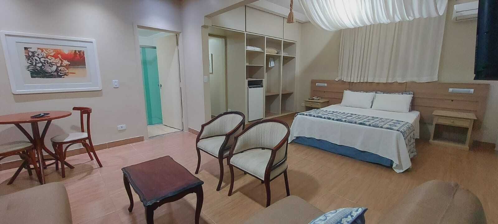 Quarto de hotel com uma cama, uma escrivaninha, duas cadeiras, uma mesinha, um guarda-roupa e um quadro na parede.