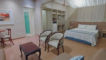 Quarto de hotel com uma cama, uma escrivaninha, duas cadeiras, uma mesinha, um guarda-roupa e um quadro na parede.